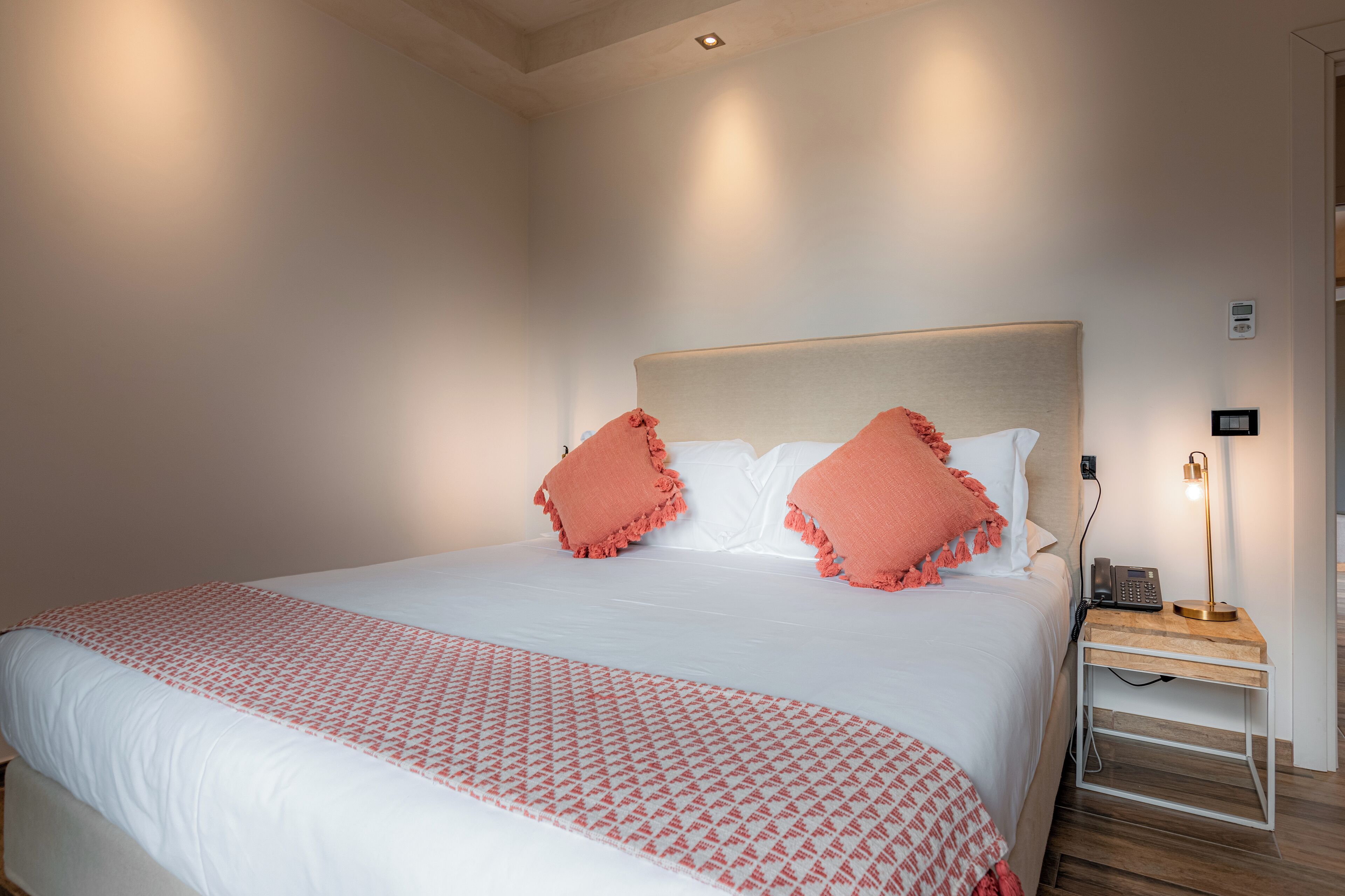 Cama bem arrumada com colcha estampada e duas almofadas, mesa de cabeceira com abajur e termostato fixado na parede.