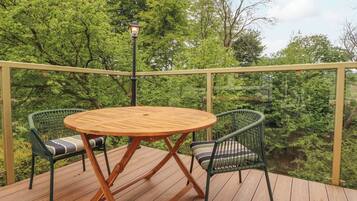 Um deck de madeira com mesa redonda e duas cadeiras, ladeado por um guarda-corpo de vidro e vista para as árvores.