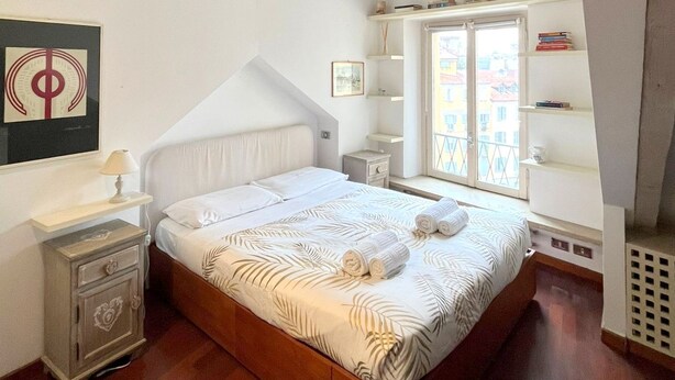 Quarto com cama de madeira, mesa de cabeceira, cômoda e janela com vista para edifícios.