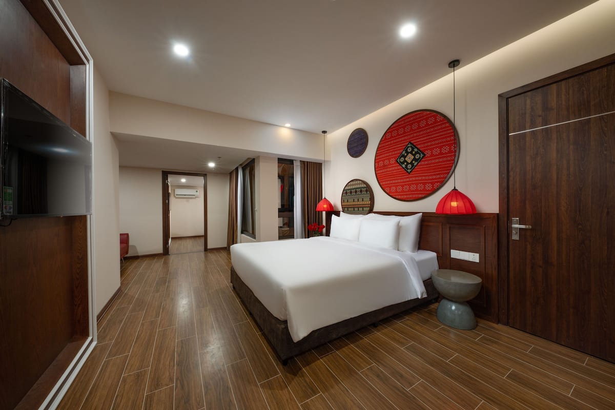 Quarto de hotel moderno com uma cama grande, piso de madeira, televisão e um detalhe decorativo vermelho na parede.