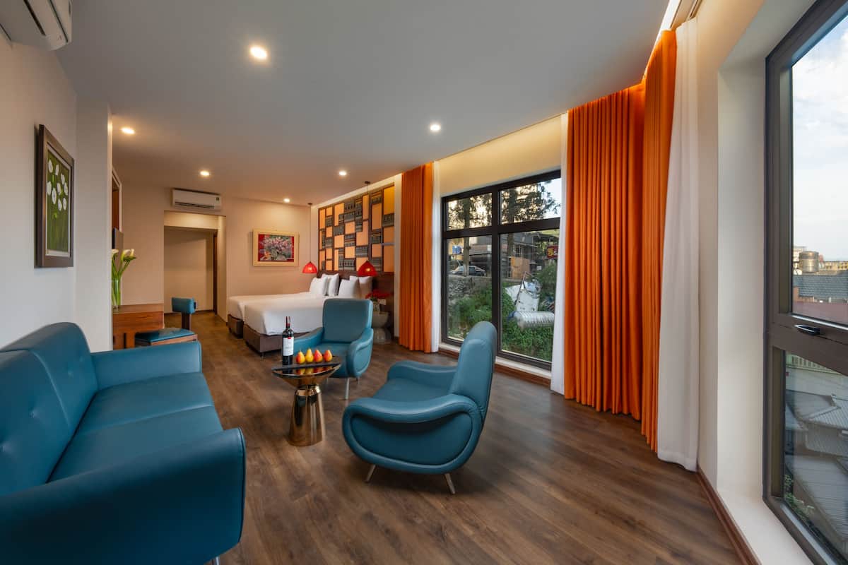 Quarto de hotel moderno com uma cama, duas poltronas azuis, uma pequena mesa de centro e uma janela grande com cortinas laranja.
