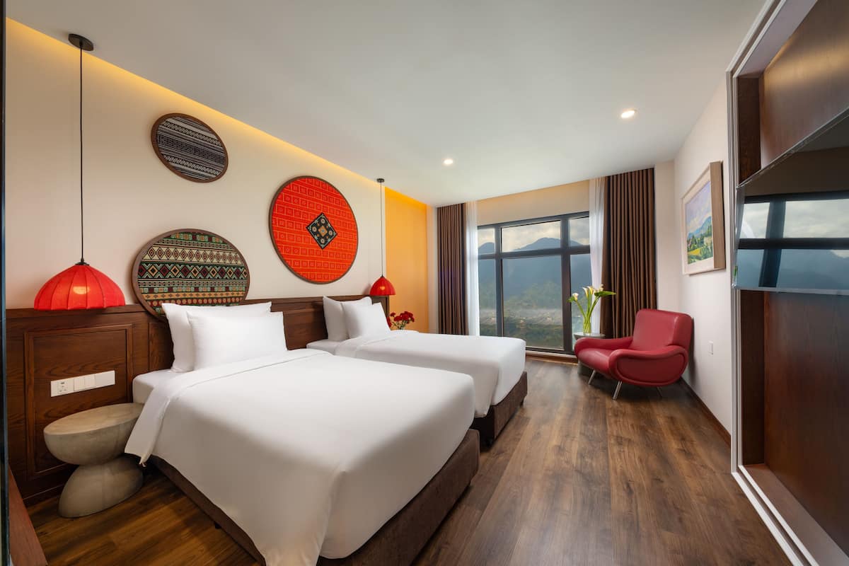 Quarto de hotel com duas camas, uma cadeira vermelha, uma janela grande com vista para as montanhas e arte decorativa na parede.