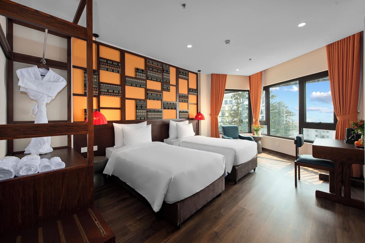 Quarto de hotel com duas camas, uma escrivaninha, uma cadeira e vista para o exterior.