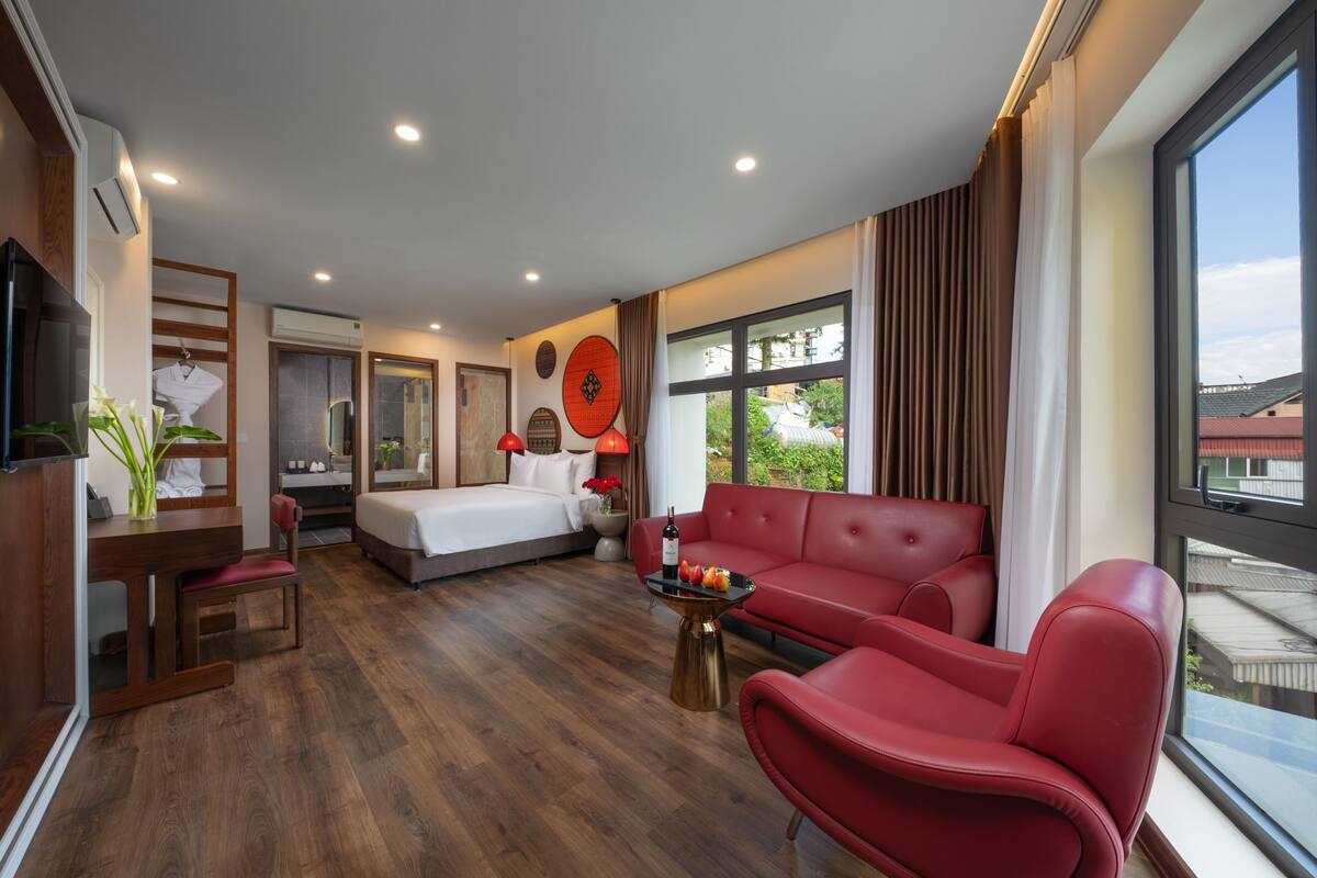 Quarto de hotel moderno com piso de madeira, uma cama, um sofá vermelho, uma mesa pequena com uma garrafa e uma televisão.
