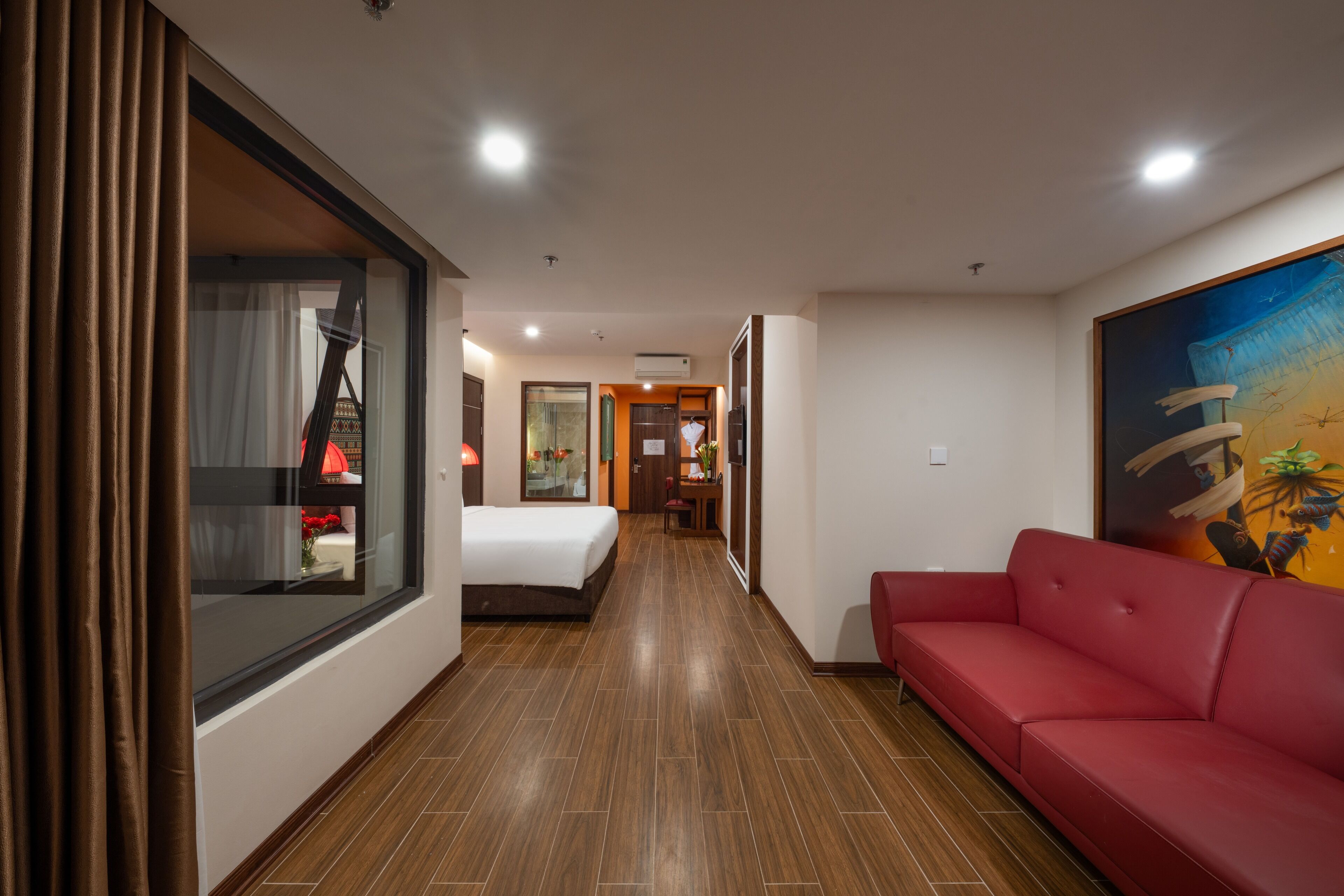 Quarto de hotel com piso de madeira, um sofá vermelho, uma cama e uma janela grande com cortinas.