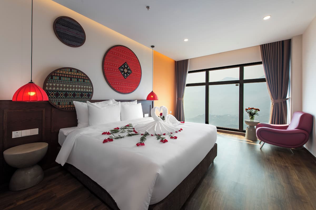 Quarto de hotel moderno com uma cama grande, um painel decorativo vermelho, uma luminária pendente vermelha, piso de madeira e vista para as montanhas pela janela.