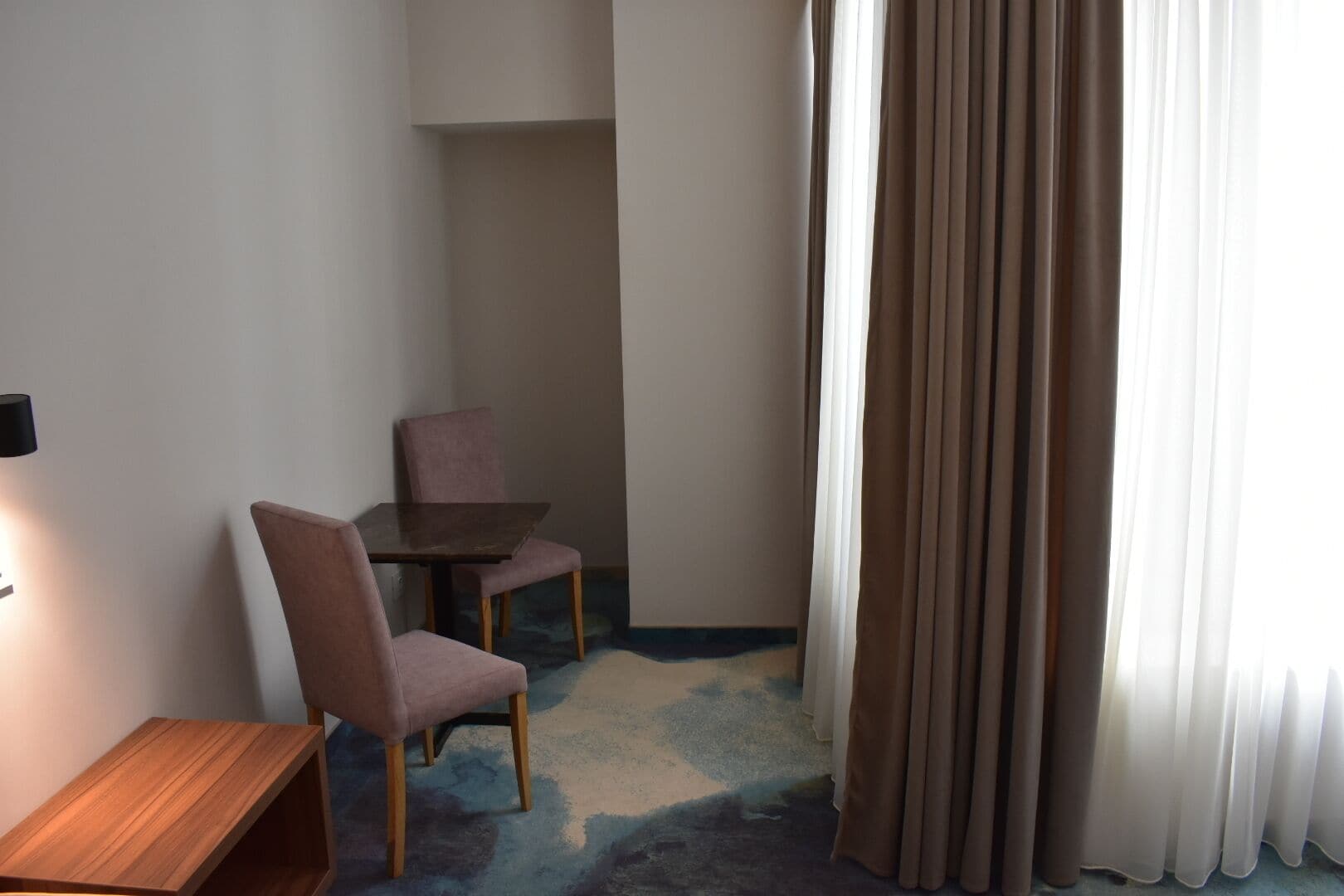 Quarto de hotel com uma pequena mesa, duas cadeiras, mesa de cabeceira de madeira e uma janela com cortinas.
