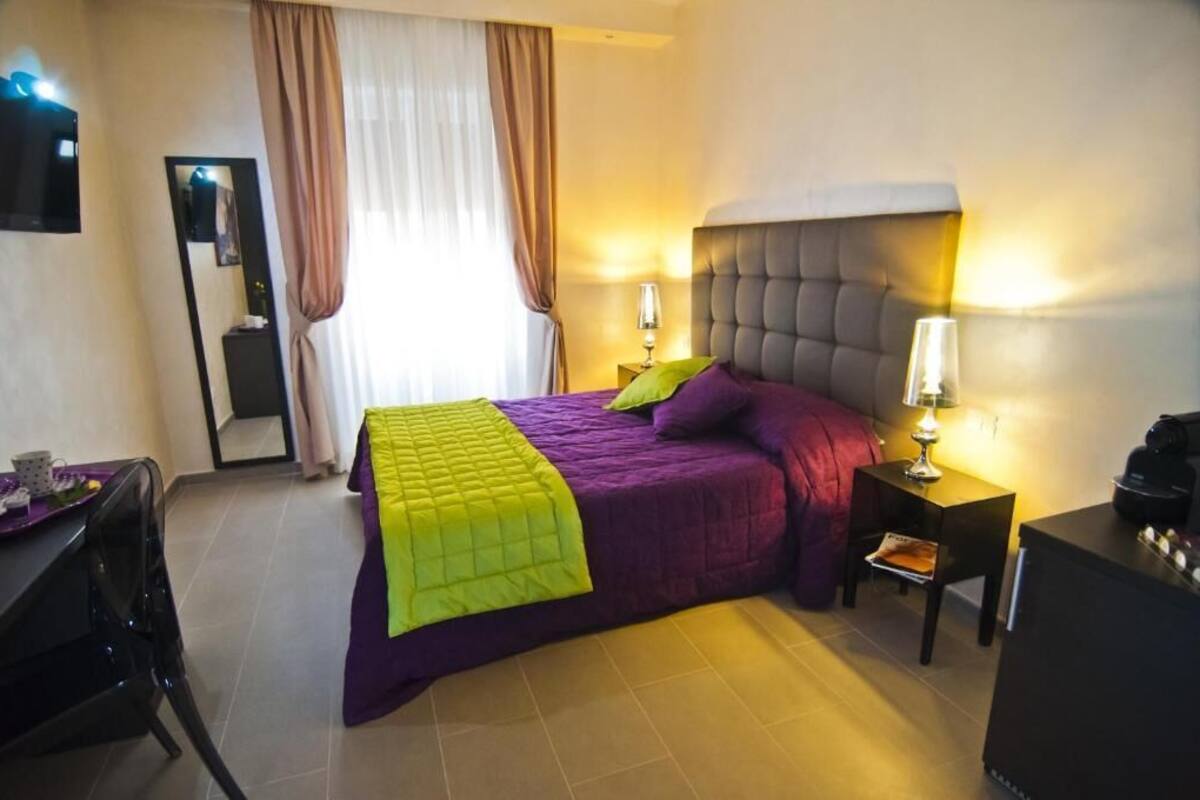 Quarto de hotel com cama, escrivaninha, cadeira e mesa de cabeceira com abajur.