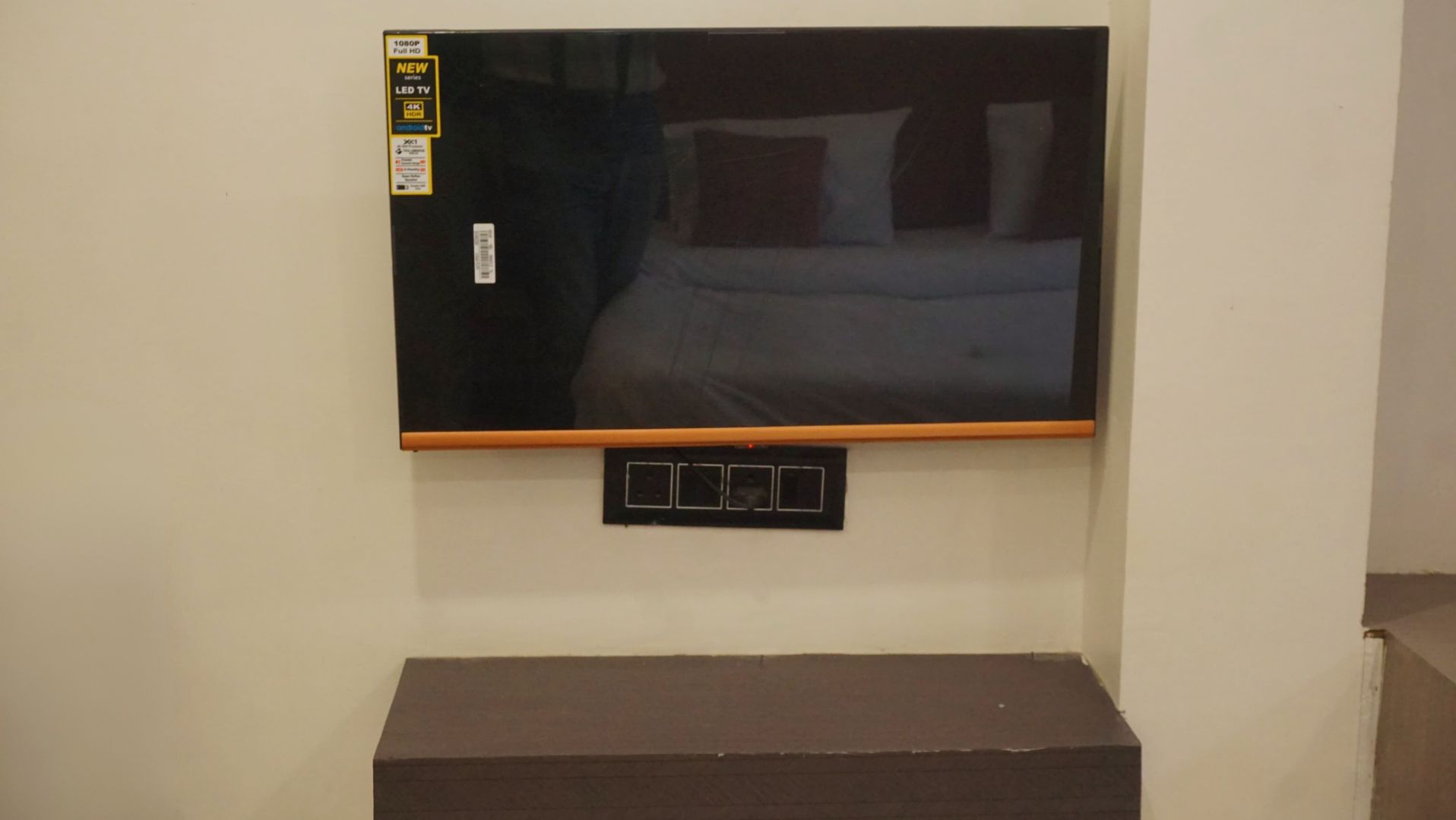 Televisão de tela plana fixada em uma parede com moldura de madeira, uma tomada elétrica abaixo e um pequeno painel de controle.