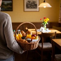 Uma cesta de vime cheia de itens de café da manhã, como croissants, frutas e suco de laranja, sendo segurada por uma pessoa vestindo um suéter cinza.