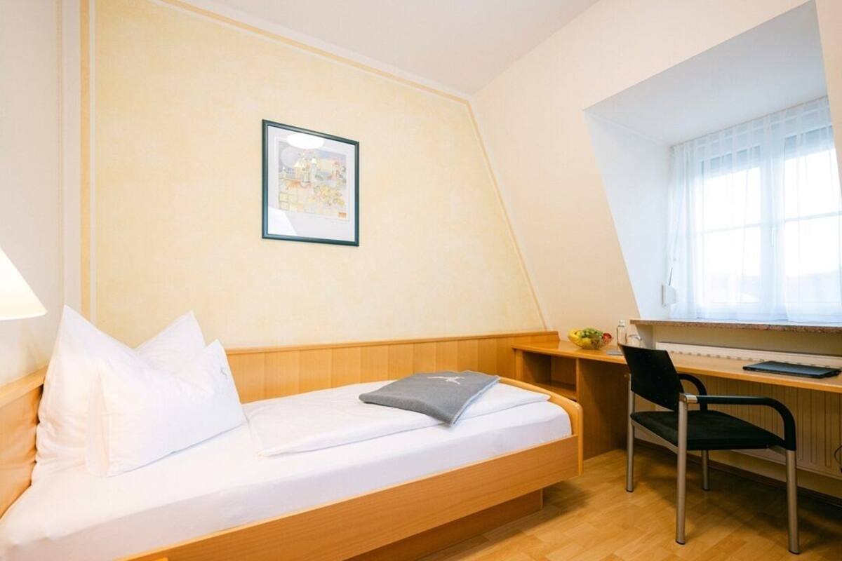 Quarto pequeno com cama de solteiro, cabeceira de madeira, escrivaninha com cadeira, janela com cortinas leves e um quadro na parede.