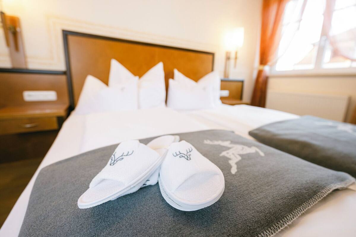 Cama bem arrumada com almofadas brancas e chinelos, manta cinza com desenho de veado, e cabeceira de madeira.