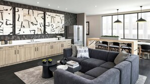 Uma sala de estar moderna com um sofá seccional cinza, uma mesa de centro preta e uma área de cozinha com armários de madeira e uma geladeira de aço inoxidável.