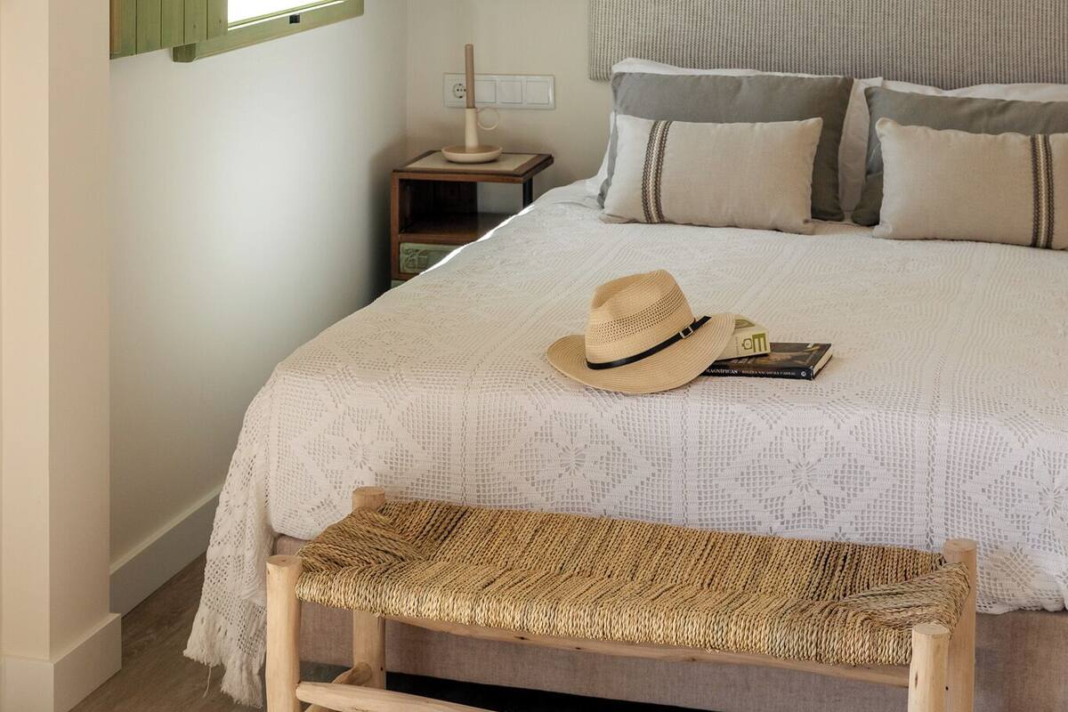 Um quarto com uma cama, uma mesa de cabeceira, um banco de vime e um chapéu sobre a cama.
