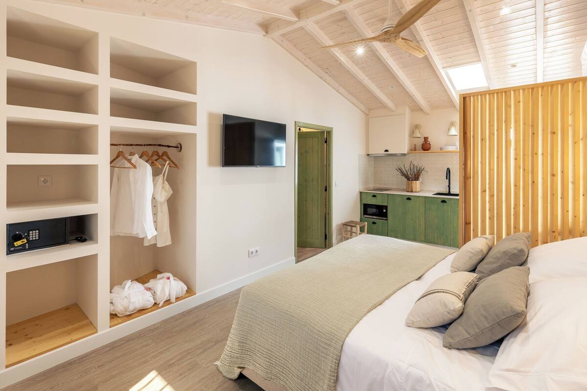 Quarto com uma cama grande, guarda-roupa com várias prateleiras, televisão e uma porta verde que dá para uma cozinha.