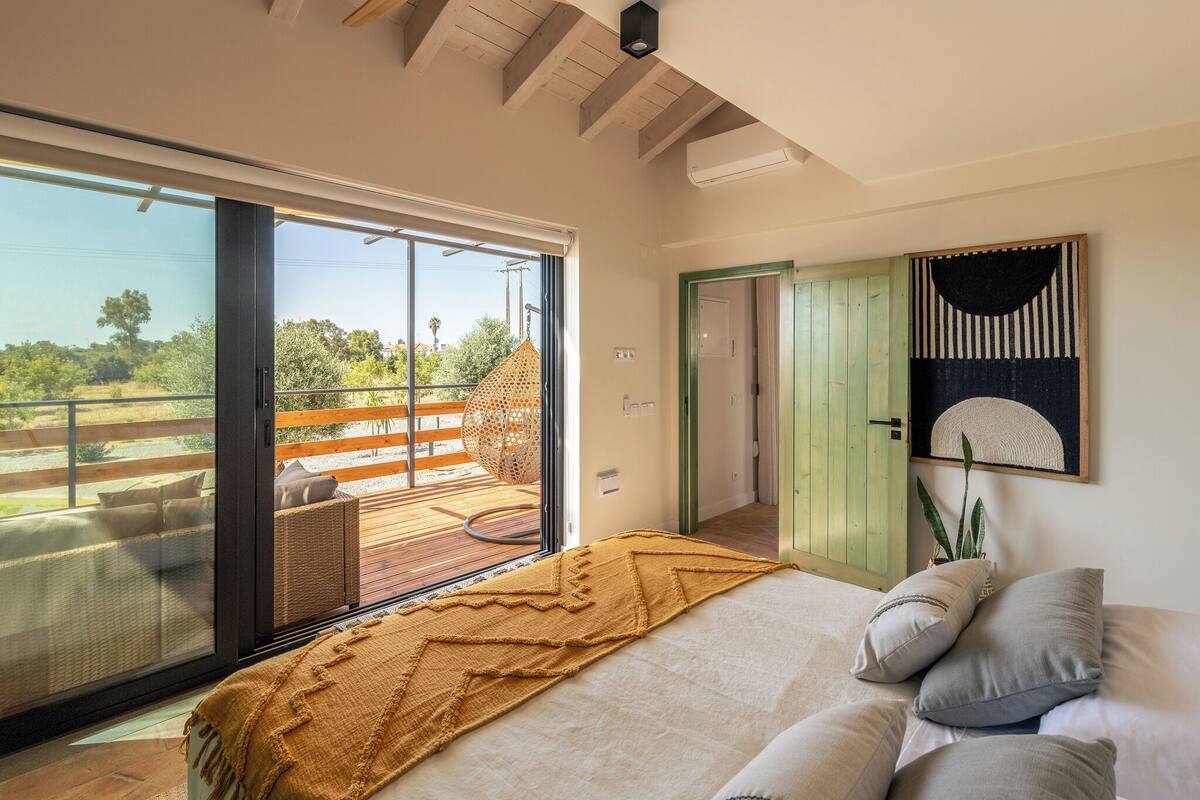 Quarto com uma cama grande, vista através de uma porta de vidro para um deque exterior, uma porta verde e uma obra de arte emoldurada na parede.