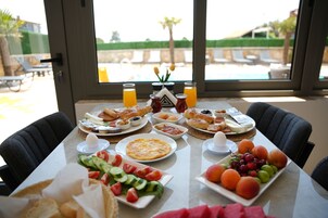 Mesa de café da manhã com diversos pratos, incluindo ovos, doces e frutas, em um quarto com grandes janelas com vista para uma área de piscina.