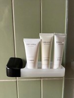 Três produtos TERRANEA de spa e bem-estar sobre uma prateleira branca, em frente a uma parede com azulejos verdes.