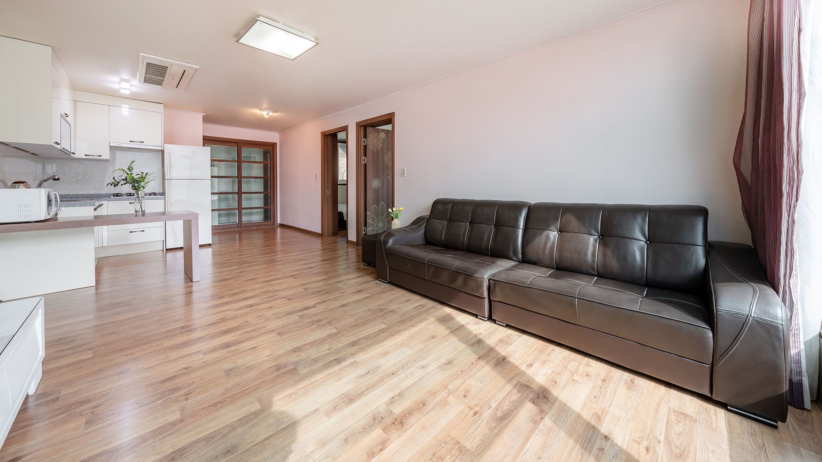 Uma sala de estar moderna com sofá de couro, piso de madeira e área de cozinha com armários brancos.