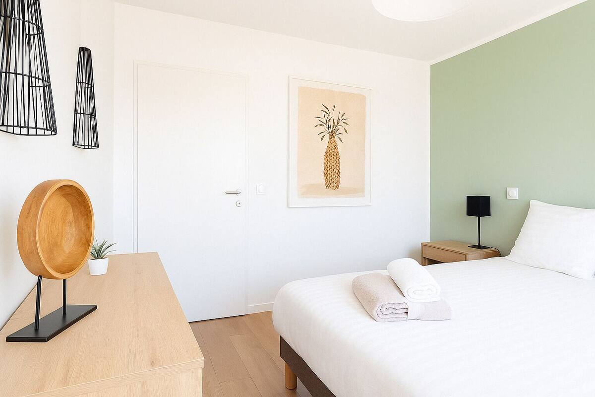 Quarto de hotel com cama, mesa de cabeceira de madeira, pequena planta em vaso, obra de arte com tema de abacaxi emoldurada e mesa de cabeceira com luminária.