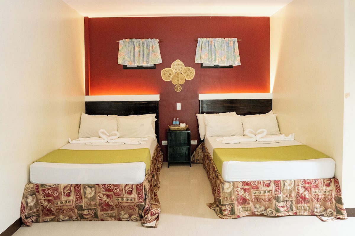 Quarto de hotel com duas camas, cada uma com colcha e almofadas estampadas. Há uma pequena mesa com uma cesta e uma garrafa entre as camas.