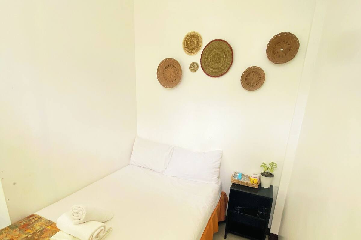 Cama bem arrumada com lençóis brancos, uma pequena mesa de cabeceira com uma planta e detalhes decorativos na parede.
