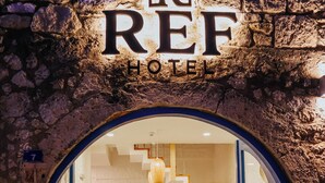 Entrada de hotel com fachada de pedra, um letreiro que diz "REF HOTEL", e um interior moderno com escadas, área de estar e plantas em vasos.