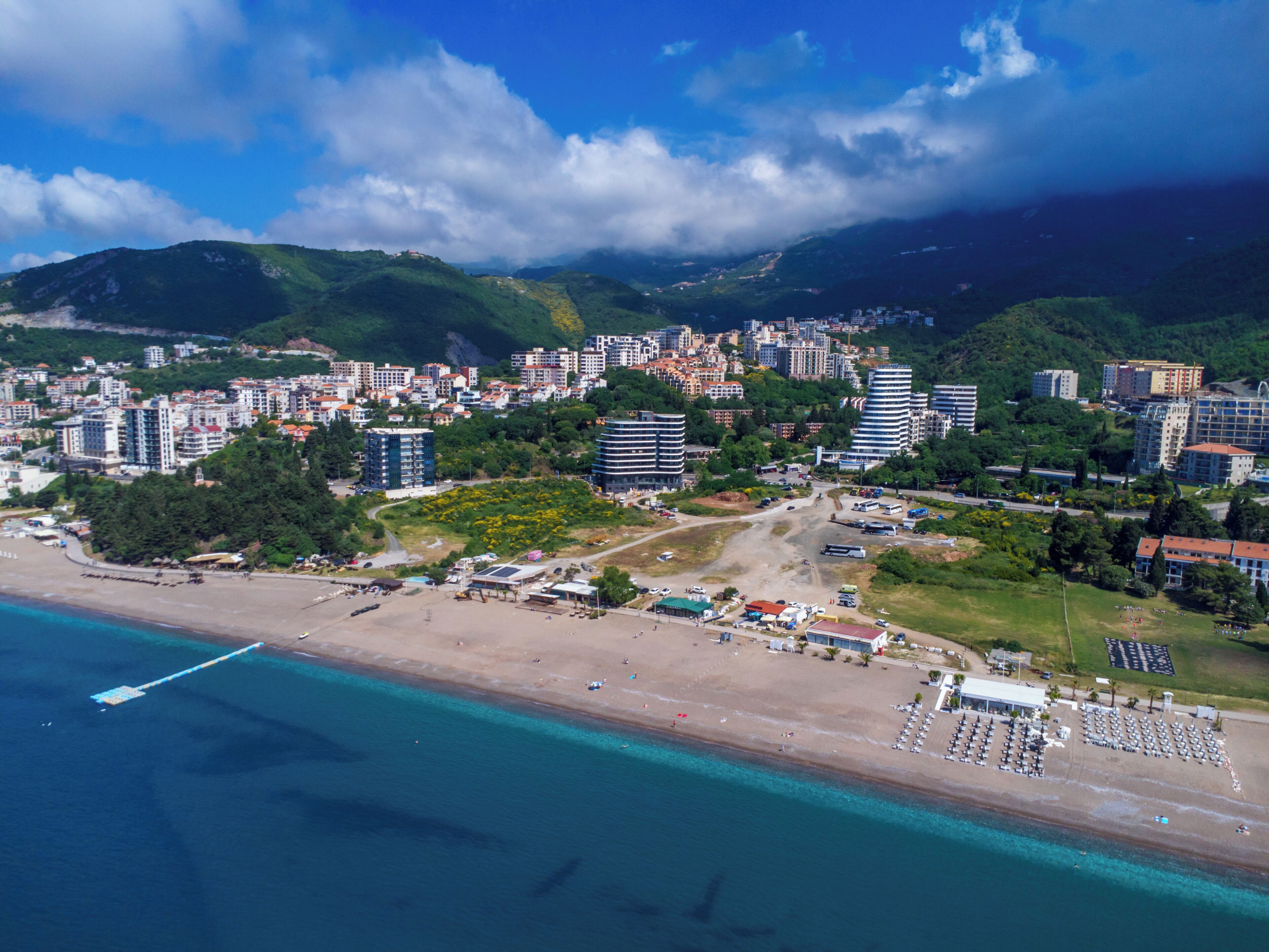 Vista aérea de uma cidade costeira com praia, edifícios e montanhas ao fundo.