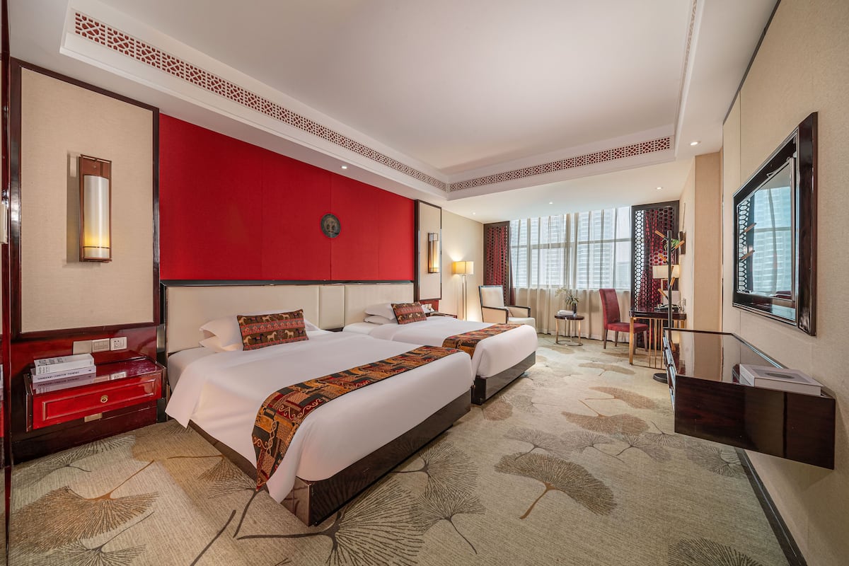 Quarto de hotel com duas camas, uma parede de destaque vermelha, uma janela grande e um balcão de recepção.