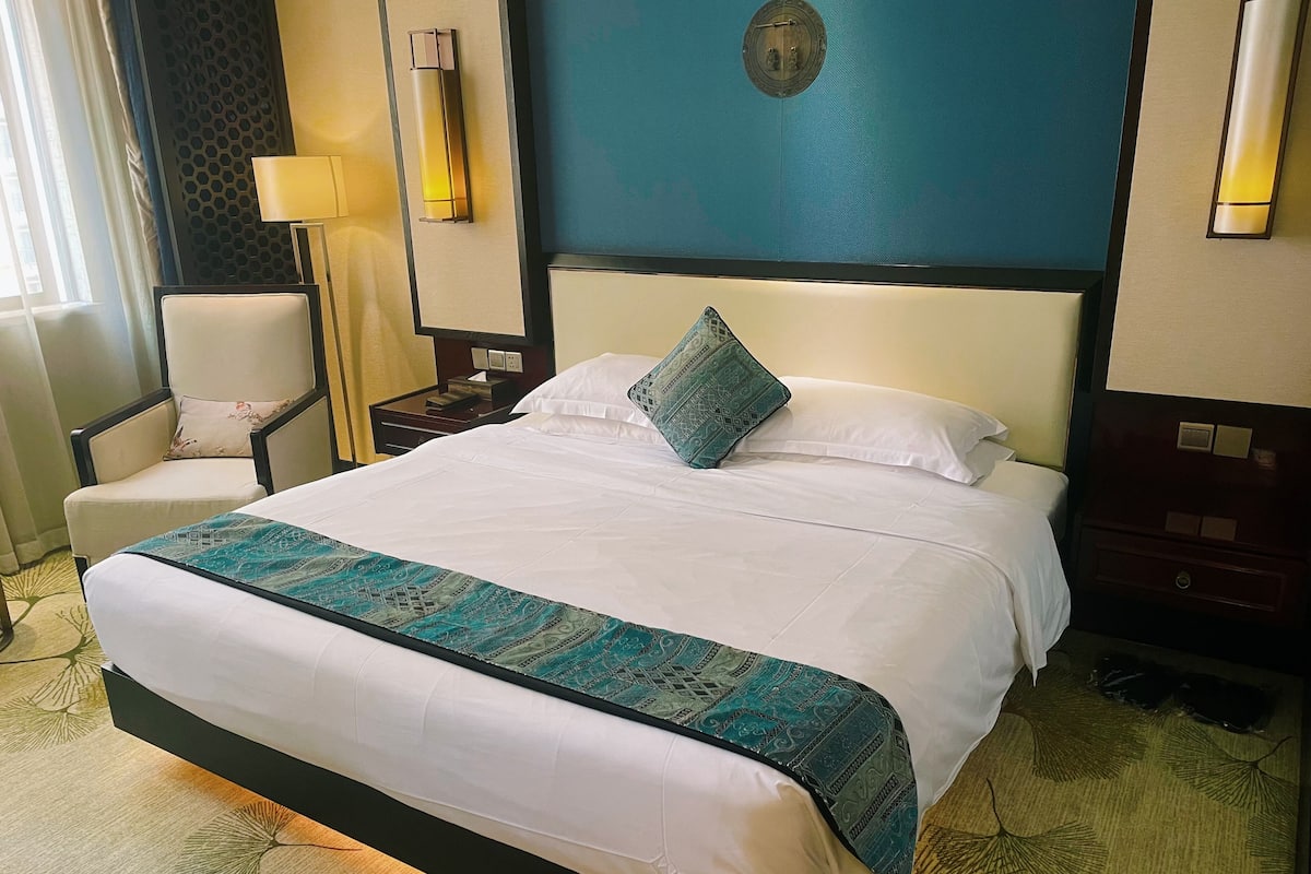 Quarto de hotel com uma cama grande, uma cadeira, um criado-mudo e um relógio de parede.