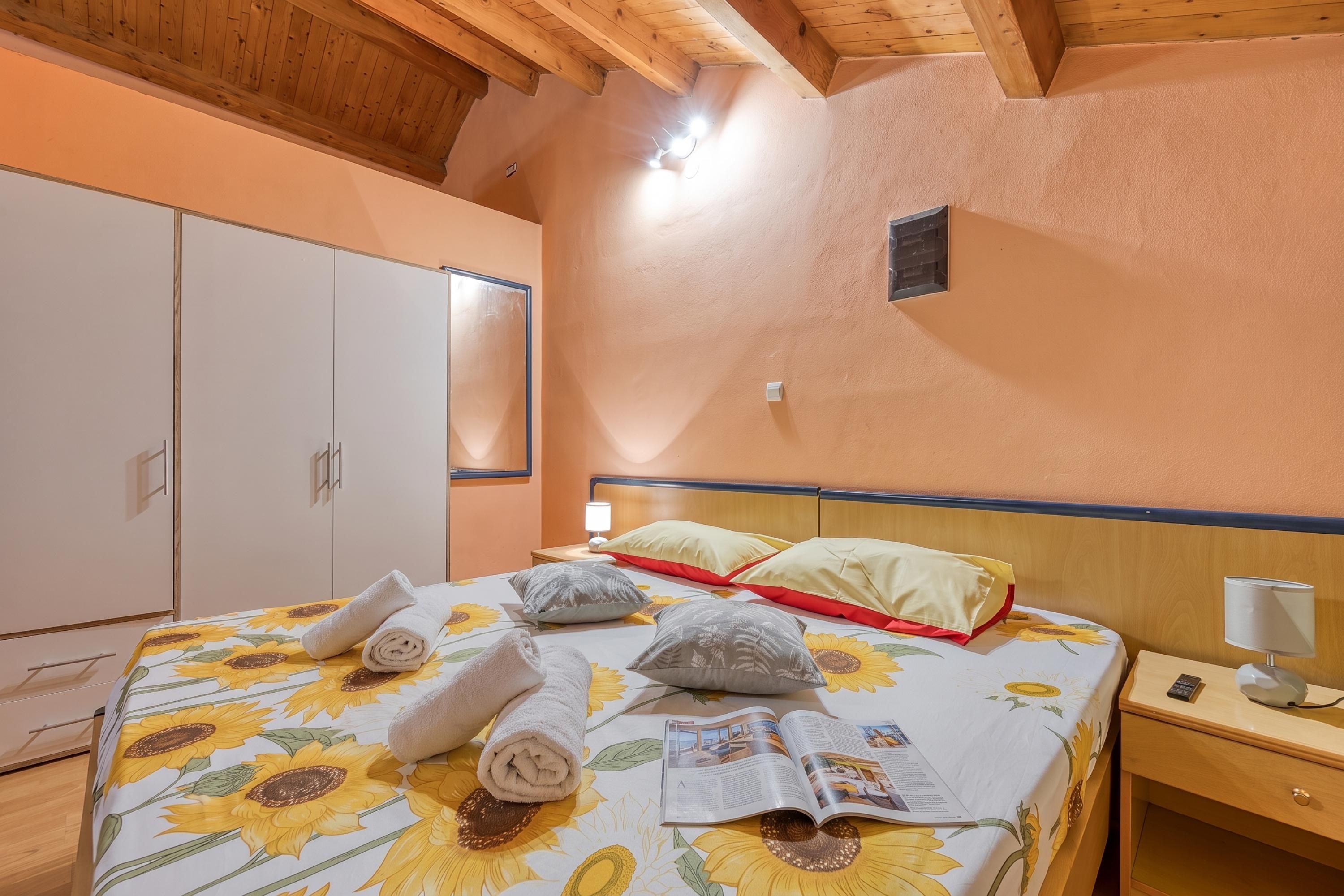 Quarto com cama, almofadas, mesa de cabeceira, luminária, livro e guarda-roupa.