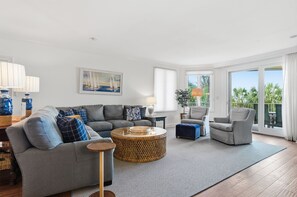 Uma sala de estar moderna com um sofá cinzento, uma mesa de centro redonda e uma poltrona cinza. Há um quadro na parede e uma janela com vista para o verde.
