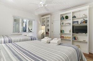Quarto com duas camas, um ventilador de teto, janela com persianas e uma estante embutida com objetos decorativos.