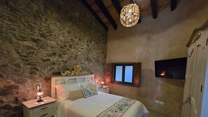Quarto com parede de pedra, cama com colcha florida, mesa de cabeceira com abajur e televisão fixada na parede.