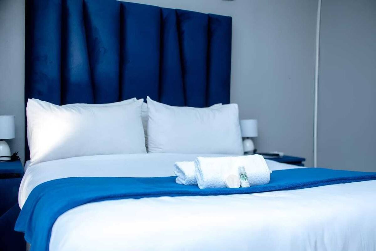 Cama bem arrumada com roupa de cama branca e azul, cabeceira azul e toalha dobrada sobre a cama.
