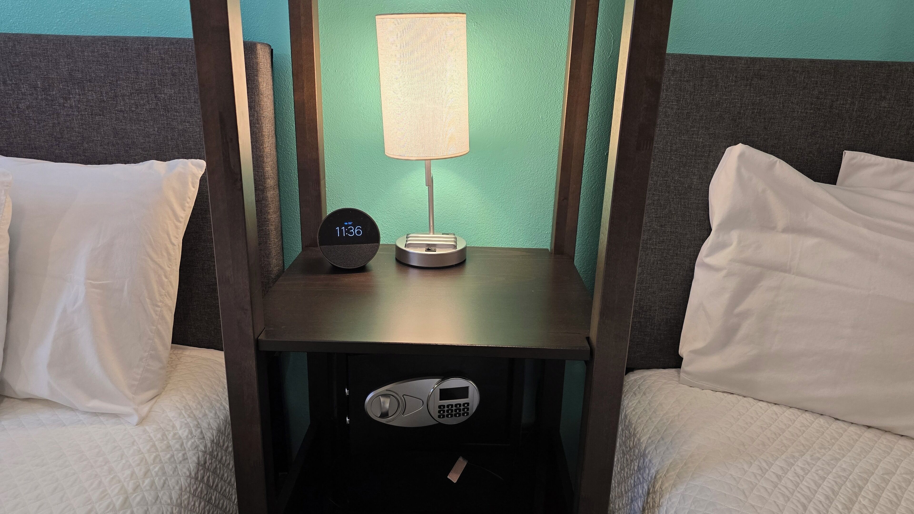 Quarto de hotel com mesa de cabeceira, luminária, relógio e uma cama.