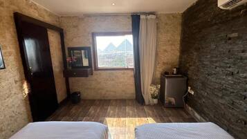 Quarto de hotel com duas camas, uma janela com vista para pirâmides, uma mesinha com uma planta e um ar-condicionado fixado na parede.