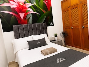 Quarto de hotel com cama, mesa de cabeceira com ventilador, criado-mudo, armário de madeira e uma grande obra de arte floral na parede.