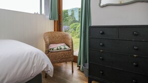 Quarto com cama, uma cadeira de vime com almofada, cômoda preta e vista para o verde pela janela.