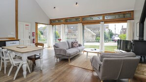 Uma sala de estar espaçosa com uma mesa de jantar de madeira, cadeiras brancas, um sofá cinza e uma pequena mesa redonda. Grandes janelas proporcionam vista para o exterior.