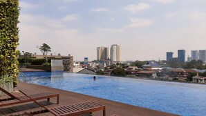 Piscina no terraço com espreguiçadeiras de madeira, vista para a cidade e um edifício com o logo 'H'.