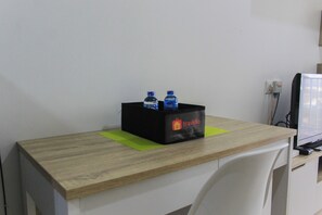 Uma pequena mesa com tampo de madeira, uma caixa preta identificada como 'travelio' contendo duas garrafas de água, e uma cadeira branca.