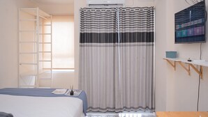 Quarto com cama, televisão na parede, janela com cortinas e prateleira com objetos.