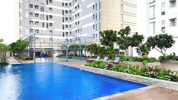 Piscina com deck de madeira, cercada por vegetação e um edifício alto ao fundo.