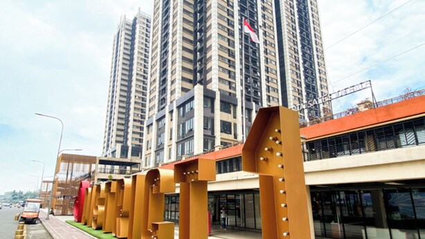 Um edifício moderno com várias estruturas douradas em primeiro plano, um arranha-céu com uma bandeira ao fundo e um céu limpo.