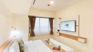 Quarto de hotel com cama, televisão na parede, janela com cortinas e mesa de cabeceira com um vaso e um folheto.