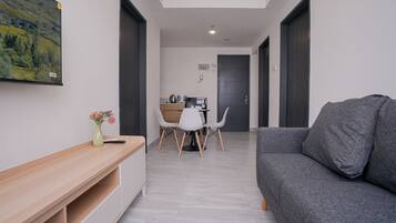 Uma sala de estar moderna com um sofá cinzento, uma mesa consola de madeira com um vaso, e uma área de refeições com uma mesa e cadeiras brancas.