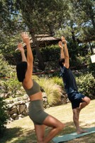 Duas pessoas praticando yoga ao ar livre em uma área gramada, com árvores e um muro de pedra ao fundo.