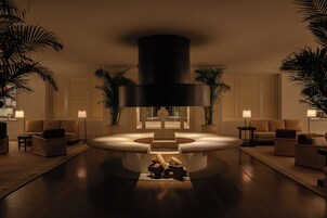 Uma sala de estar moderna com uma área de estar circular central, rodeada por sofás e luminárias, e uma grande estrutura cilíndrica acima.
