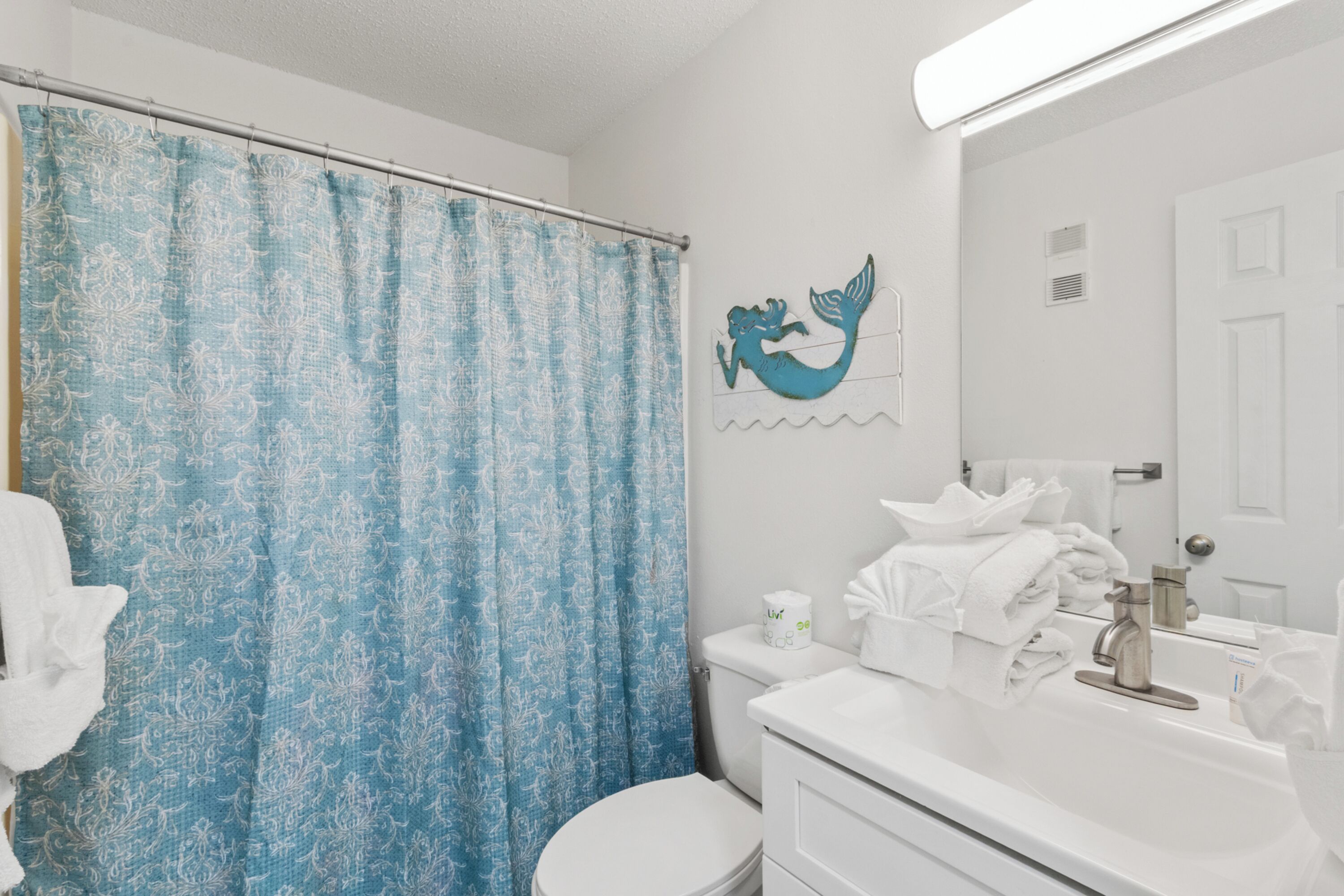 Casa de banho com sanita branca, lavatório com espelho e cortina de chuveiro azul.
